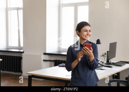 Happy freelance worker woman holding smartphone, sitting at work table ...