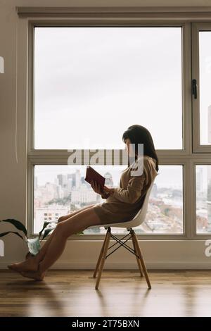 Woman reading a book at big window with city view. Young female ...