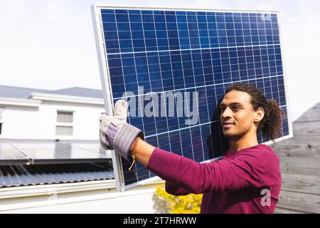 Happy biracial man carrying solar panel and smiling in sunny garden ...