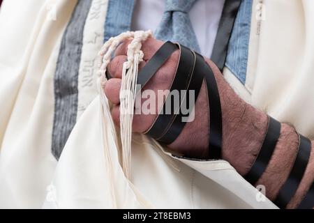 A hand gathers and holds the tzitzit, strings tied to the four corners ...