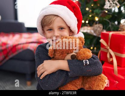 Adorable toddler hugging teddy bear standing by christmas tree at home ...