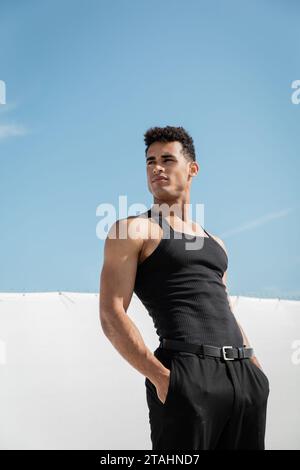 muscular young cuban man posing near white wall with shadow on urban ...