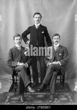 Edwardian formal studio portrait of three men in suits, two sitting and ...