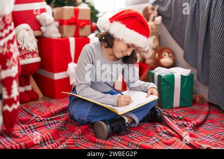 Adorable chinese girl writing on notebook sitting on floor by christmas ...