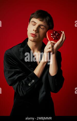 sentimental man in velvet blazer holding heart-shaped gift box on red ...