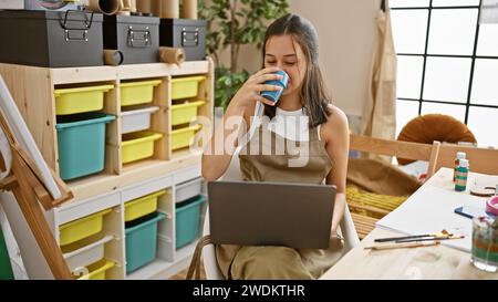 Young hispanic woman artist drinking coffee sitting on floor at art ...