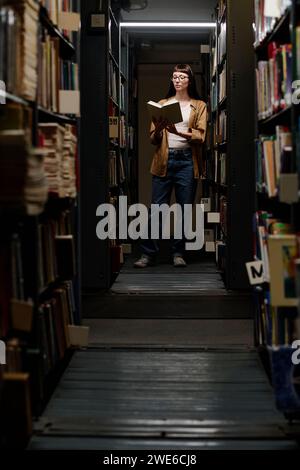 Student reading book and standing amidst shelves in library Stock Photo ...