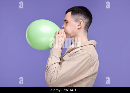 Young man inflating light green balloon on purple background. Space for ...