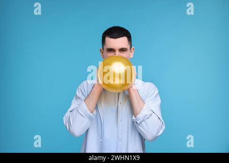 Young man inflating golden balloon on light blue background Stock Photo ...