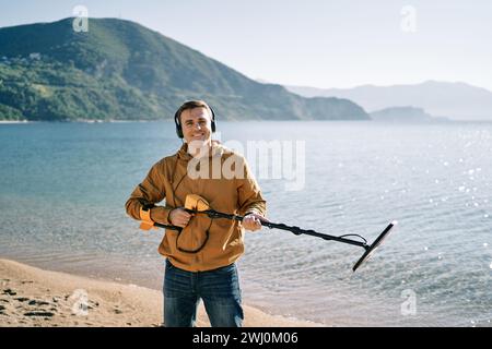 Smiling man with a metal detector at the ready stands on the seashore ...