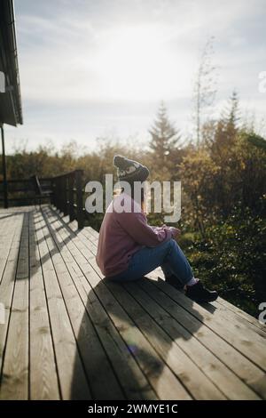 Peaceful woman sitting on wooden cabin balcony at dusk holds cup ...