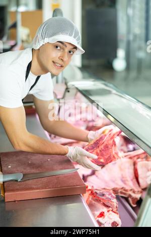 Positive young salesman demonstrating piece of meat in butcher shop ...