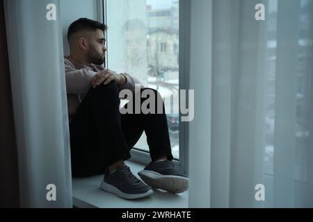 Sad man sitting on sill and looking at window indoors Stock Photo - Alamy