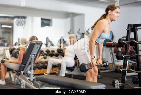 Slender girl trains muscles of arms and back using barbell and ...