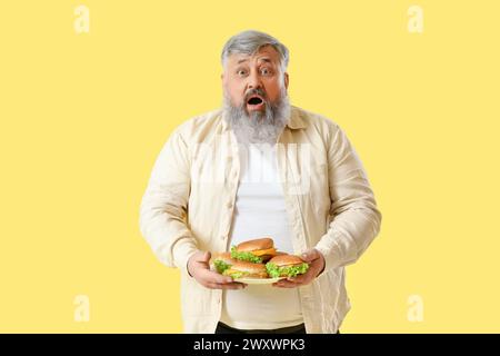 Overweight mature man with burgers on yellow background. Overeating ...