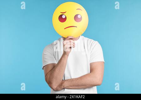 Man covering face with thinking emoticon on light blue background Stock ...