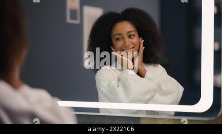 African girl in bathrobe is looking in the mirror on her face in white ...