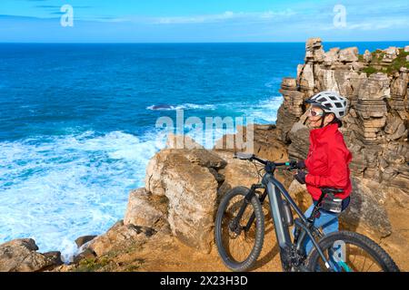 Brave senior woman riding her electric mountain bike on the rocky cliffs of Peniche at the ...