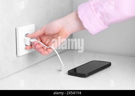 Woman plugging smartphone into power socket at white table indoors ...