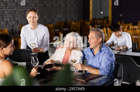 Waitress bringing aperitif to elderly couple selecting dishes from menu ...