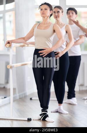 Female ballet teacher exhibiting third position at barre during group ...