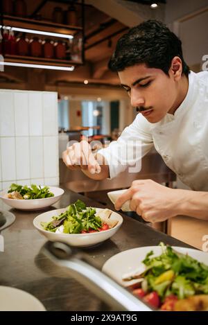 Focused male chef sprinkling salt on salad while bending in commercial ...