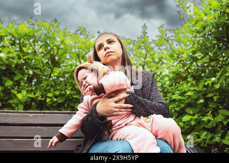 A tired mother sits on a park bench with her baby who is crying ...