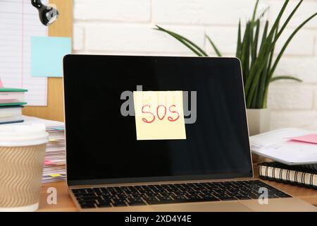 Note with word SOS, laptop and stationery on wooden table at workplace ...