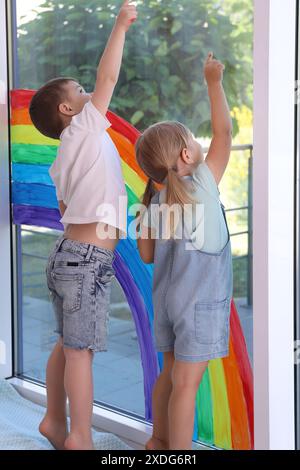 Little girl touching picture of rainbow on window indoors, back view ...