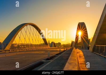 Modern arch bridge captured during sunset, with warm sunlight peeking ...
