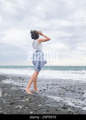 Happy woman greets stormy sea on pebble beach. Woman with hair ruffled ...
