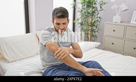 Hispanic man applying lotion to elbow in a bedroom setting, depicting ...