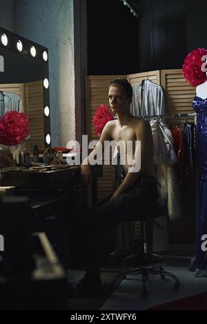 A drag queen sits at a dressing table, leaning on their hand, and gazes ...