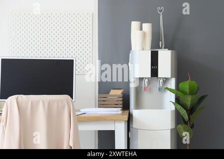Modern water cooler with paper cups on table and shelf unit near blue ...