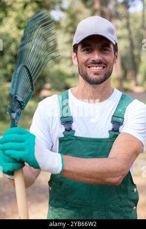 Portrait of smiling gardener carrying rake on shoulders at plant ...