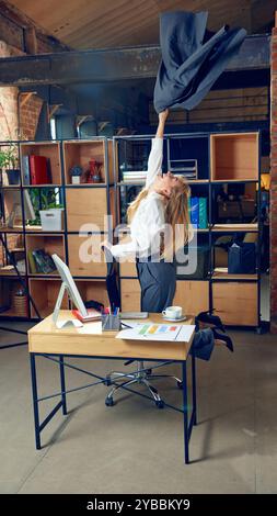 Emotional woman dancing her desk, throwing her jacket into air, smiling ...