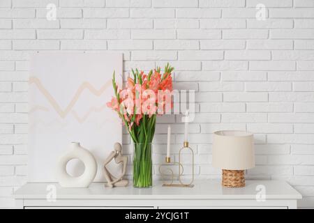 Vase with gladiolus flowers, lamp and decor on commode near white brick ...