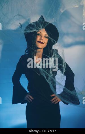 A charming witch poses in front of spider webs, her hands raised in a ...