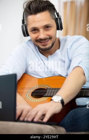 Man taking online guitar lessons with a laptop at home sitting on the ...