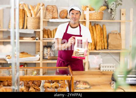 Middle-aged salesman offering a piece of cake packed in box in bakery ...