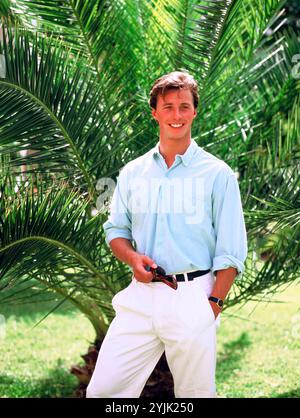 Young man standing outdoors in a park, smiling and looking away ...