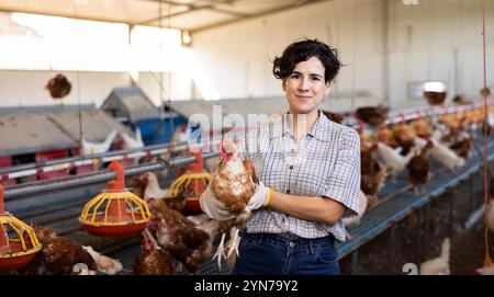 Positive young Latin female farmer holding chicken and cheking health ...