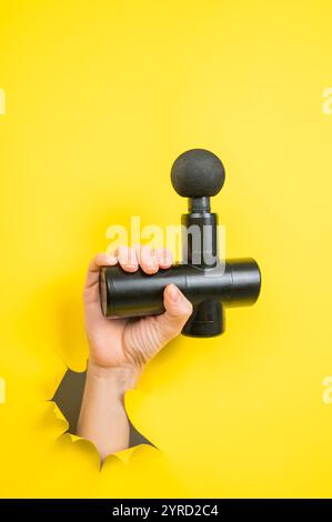 Female hand sticking out with massage gun from yellow paper background ...