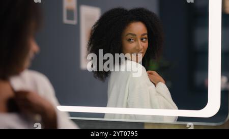 African girl in bathrobe is looking in the mirror on her face in white ...