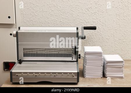 Modern binding machine and stacks of notebooks on wooden table indoors ...