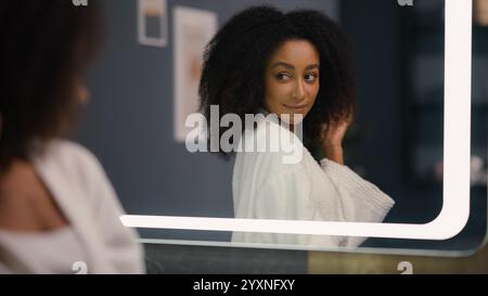 African girl in bathrobe is looking in the mirror on her face in white ...