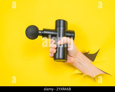 Female hand sticking out with massage gun from yellow paper background ...