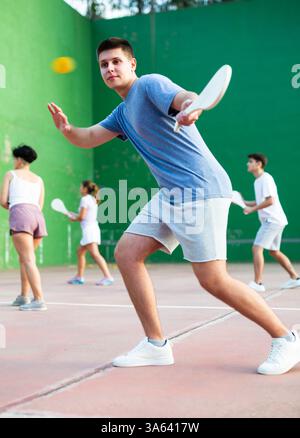 Sporty guy playing pelota with wooden racket on open fronton court ...