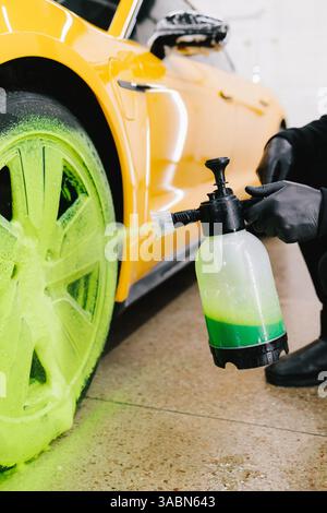 Worker cleans the tire of a yellow car using a spray with soap solution ...