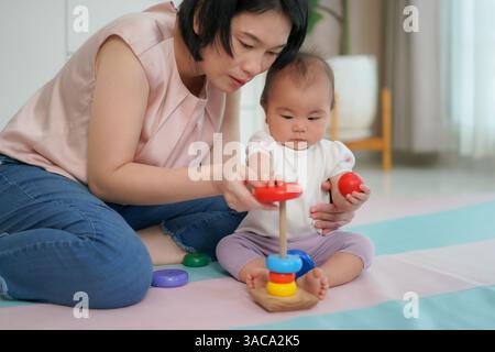 A mother guiding her baby to stack colorful wooden rings on a peg toy ...